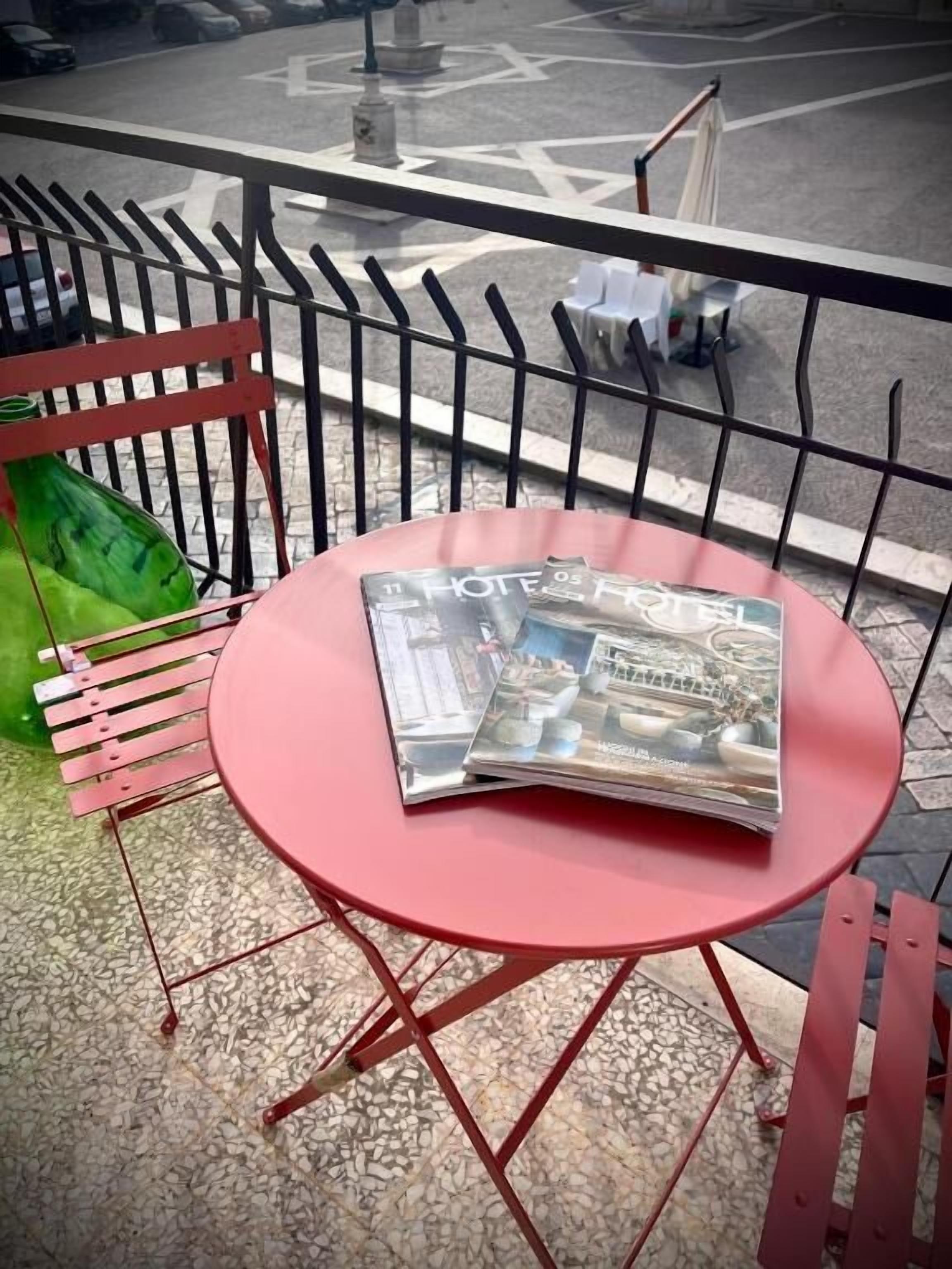 Uma mesa vermelha com uma revista, uma garrafa verde e uma cadeira vermelha em uma varanda com guarda-corpo de metal.