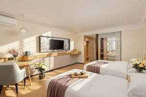 Quarto de hotel moderno com televisão de tela plana, duas camas, uma escrivaninha com cadeira e uma janela grande.