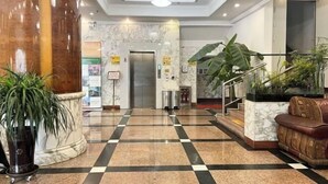 Um hall de hotel com piso xadrez, paredes de mármore, balcão de recepção, escada e plantas em vasos.