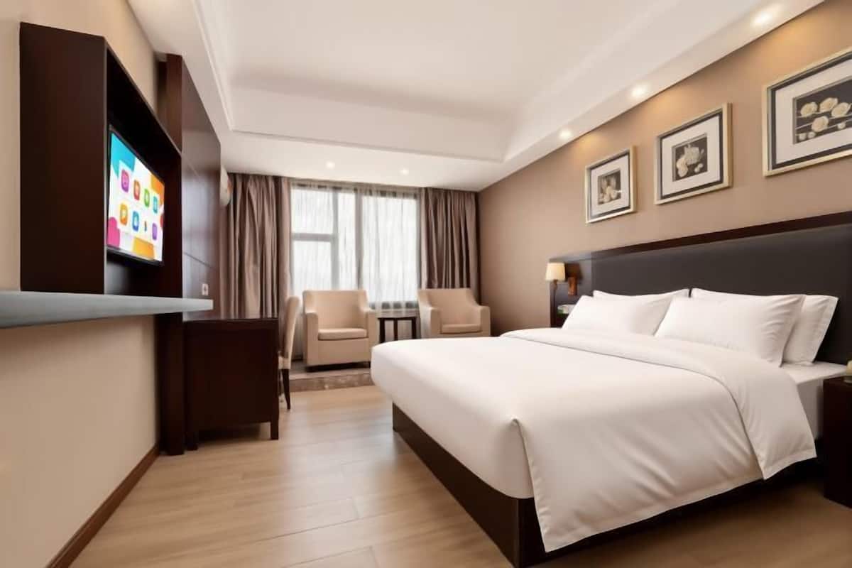 Quarto de hotel com uma cama grande, televisão presa à parede, escrivaninha com cadeiras e janela com cortinas.