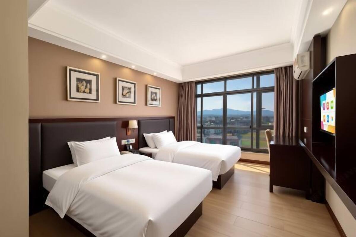 Quarto de hotel com duas camas, uma janela grande com vista para a cidade e uma televisão à direita.