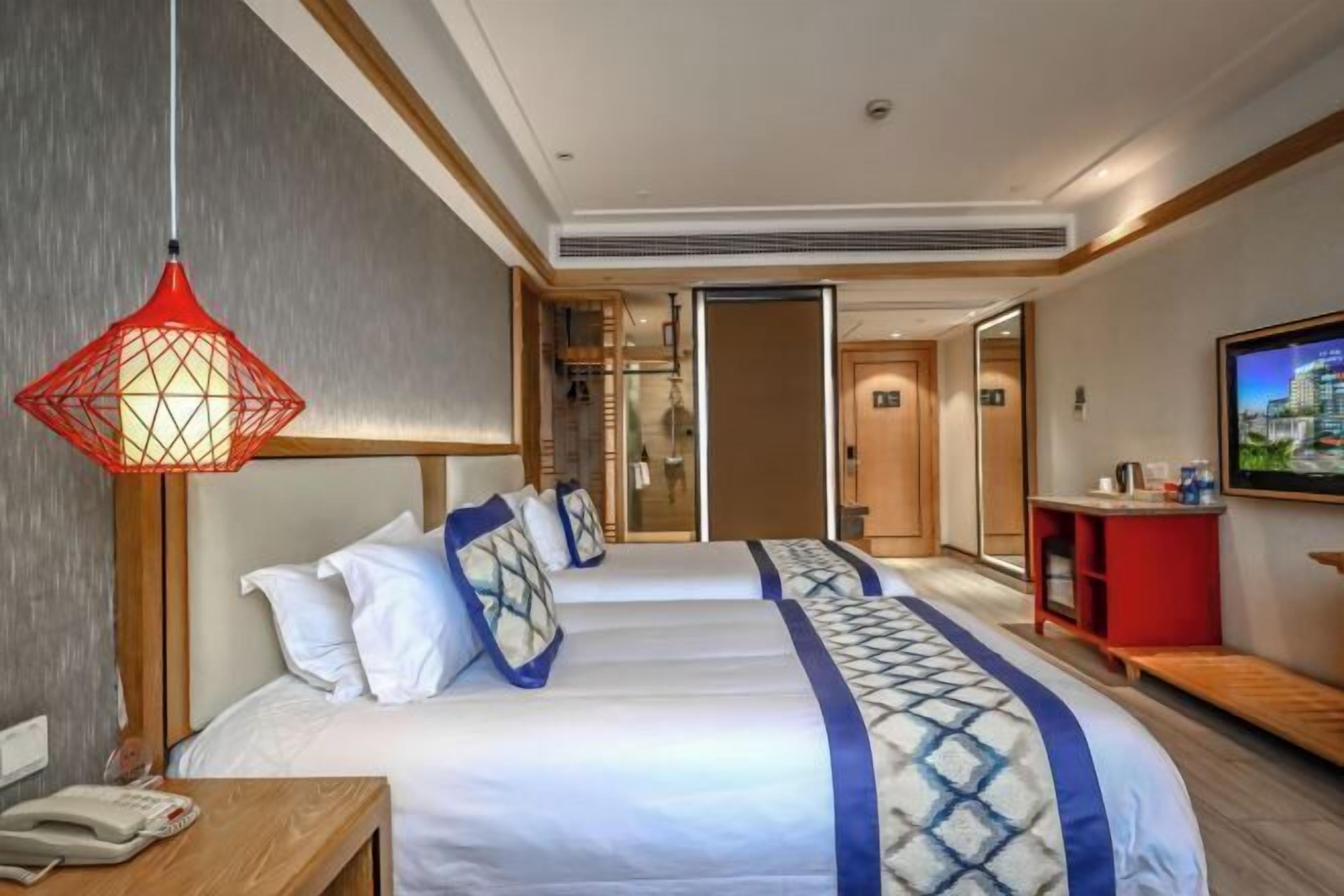Quarto de hotel com uma cama grande, uma escrivaninha com telefone, um candeeiro pendente vermelho e uma televisão na parede.