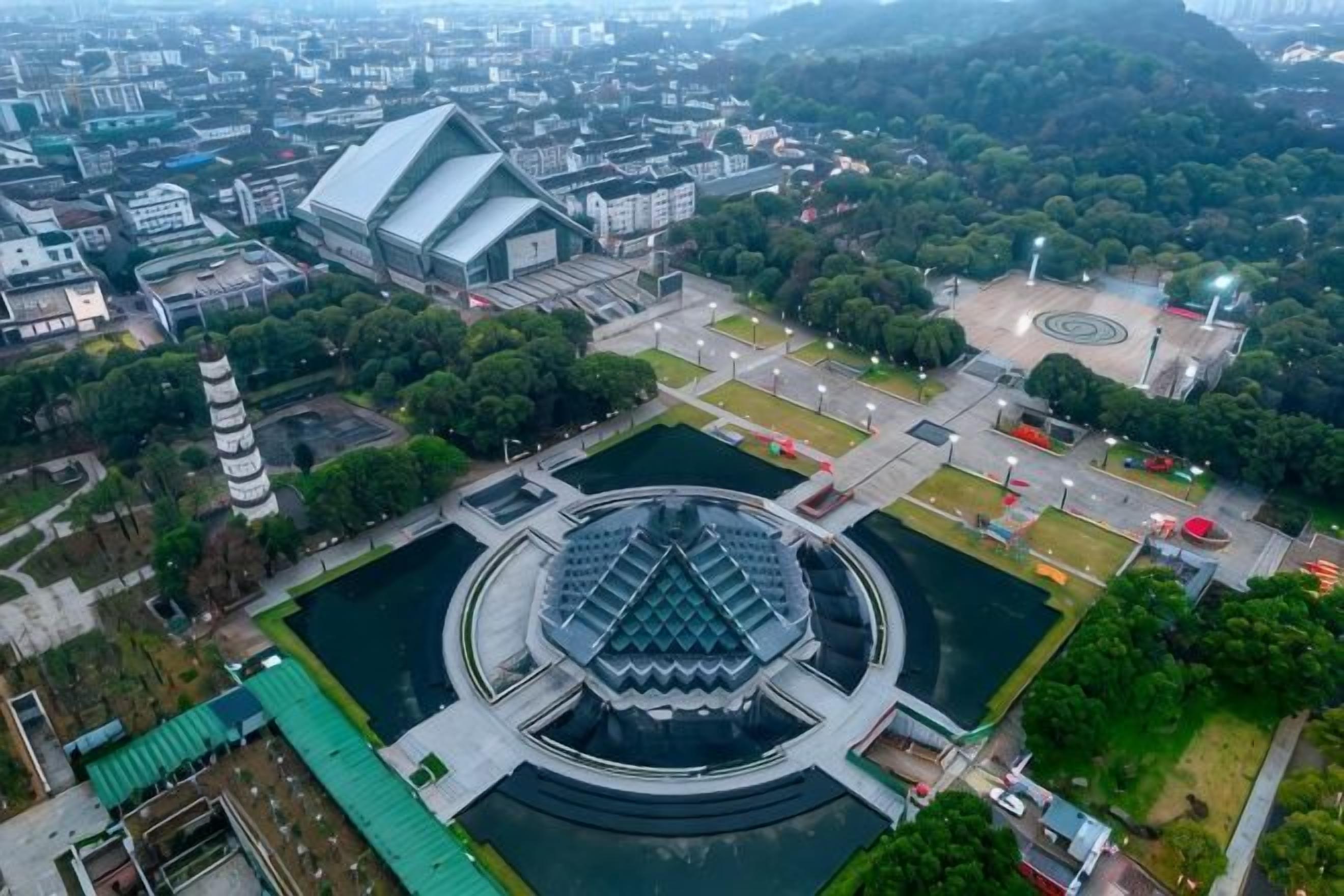 Vista aérea de uma paisagem urbana com um edifício circular imponente, cercado por água, uma mesquita com um minarete e um estádio grande.