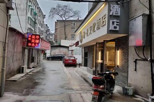 Uma rua estreita com um hotel à direita, um carro vermelho estacionado em frente e uma motocicleta na calçada.