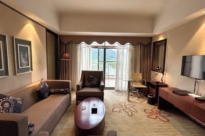 Quarto de hotel moderno com sofá, poltrona, mesa de centro redonda e uma TV.