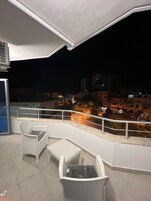 Uma varanda com móveis de vime branco, uma porta de vidro e vista para a cidade à noite.