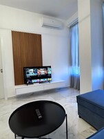 Uma sala de estar moderna com uma TV de tela plana fixada em uma parede branca, uma mesa redonda preta com um controle remoto, um sofá azul e uma janela com cortinas.