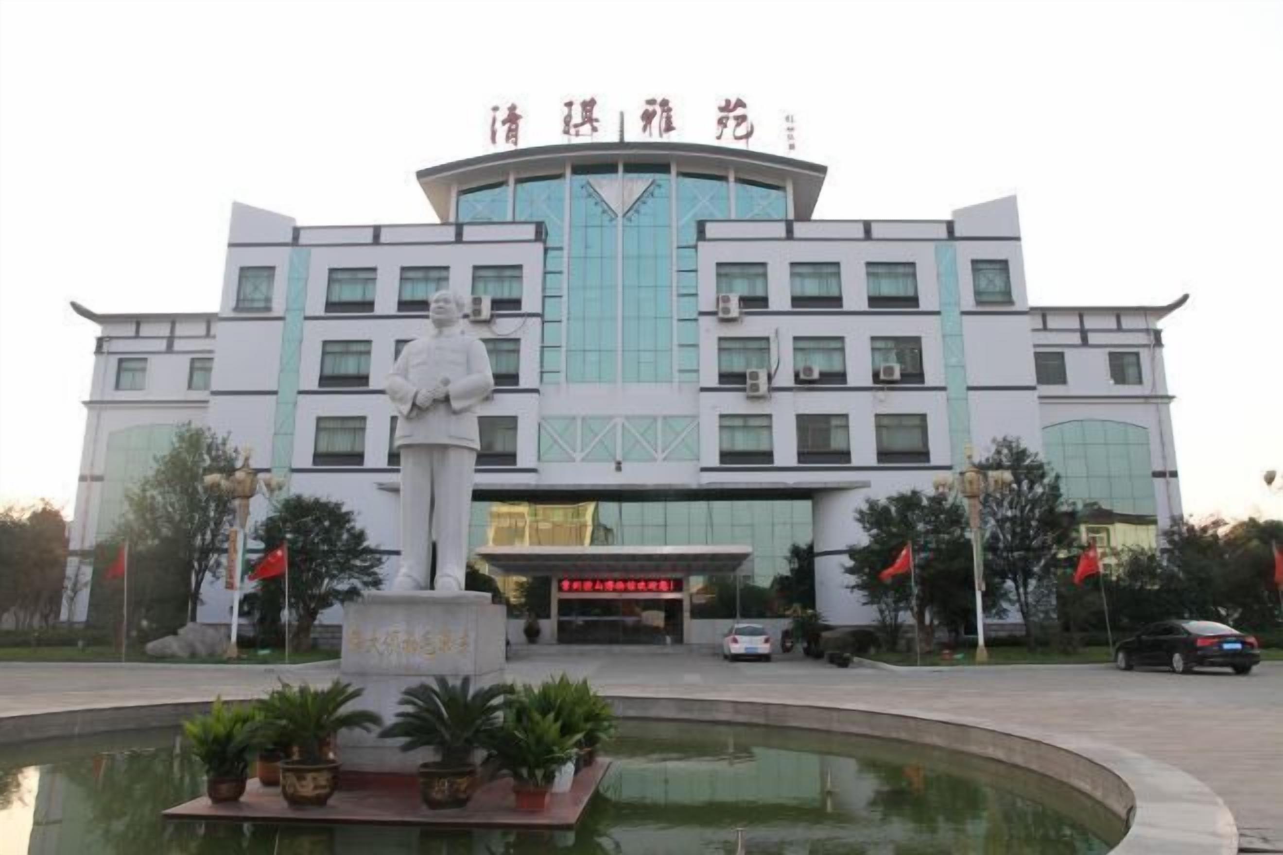 Um edifício moderno com entrada central, uma estátua em frente e uma pequena piscina com plantas em vasos.