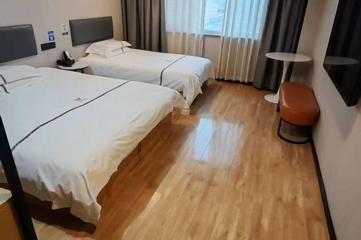 Quarto de hotel com duas camas, uma pequena mesa, um banco, mesa de cabeceira e janela com cortinas.