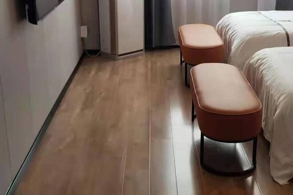Quarto de hotel com piso de madeira, dois pufes bege, cama com roupa de cama branca e uma TV fixada na parede.