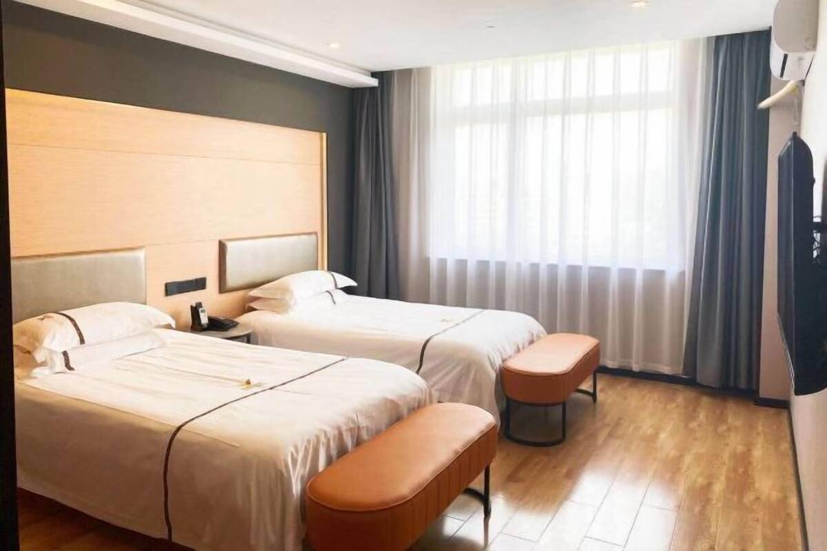 Quarto de hotel com duas camas, um banquinho, uma mesa de cabeceira, um telefone e uma janela com cortinas.