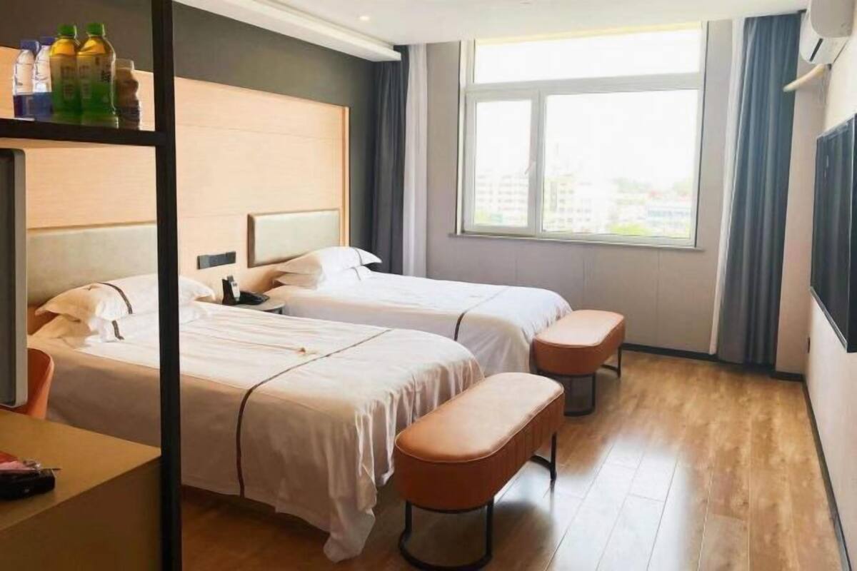 Quarto de hotel com duas camas, um banquinho, uma escrivaninha e janela com vista para a cidade.