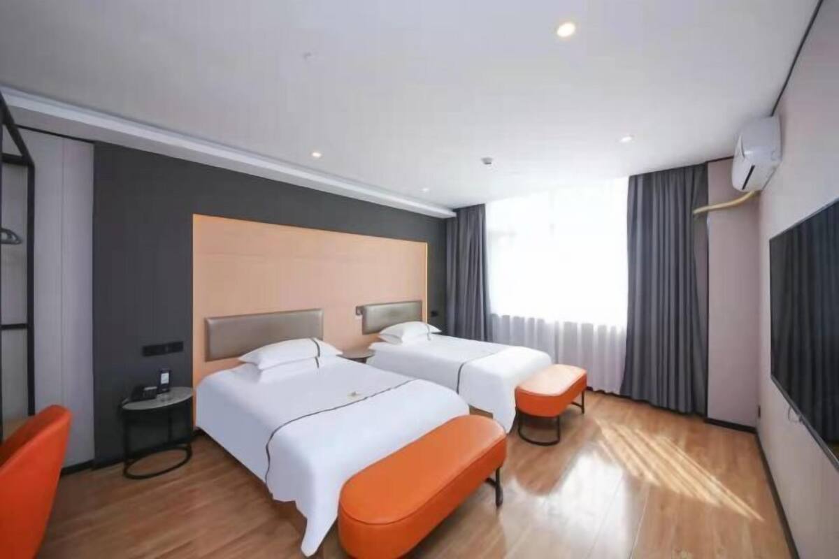 Quarto de hotel com duas camas, uma mesinha, um banco laranja e uma janela grande com cortinas.