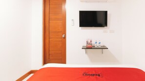 Quarto de hotel com porta de madeira, televisão montada, prateleira pequena com garrafas de água e colcha vermelha.