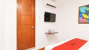 Quarto de hotel com porta de madeira, ar condicionado, televisão, prateleira pequena com garrafas de água e uma cama com manta vermelha.