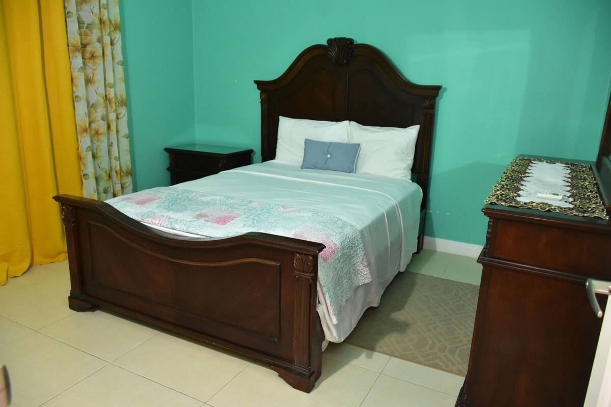 Quarto com cama de madeira, mesa de cabeceira, cômoda e cortina.