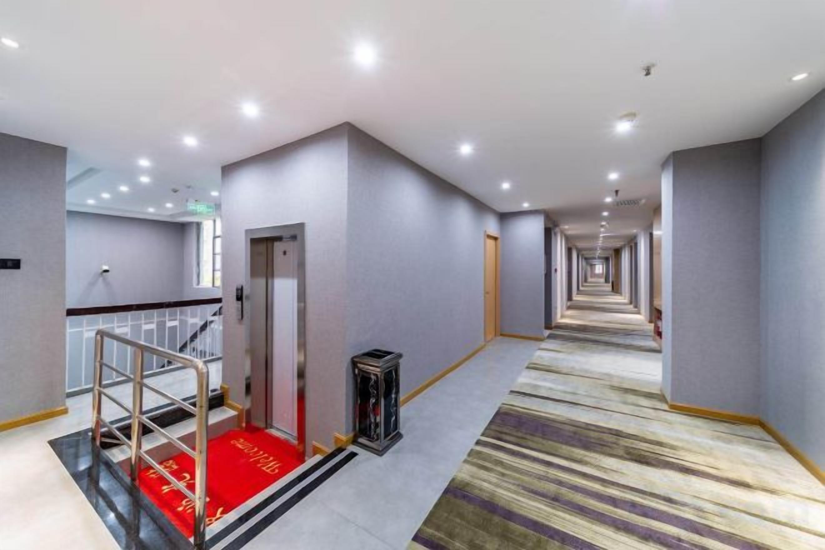 Corredor de hotel com tapete vermelho, corrimão de metal e piso coberto por carpete.