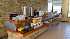 Um buffet de café da manhã com diversos alimentos e bebidas, incluindo máquinas de cereal, cafeteiras e um dispensador de ponche de frutas.