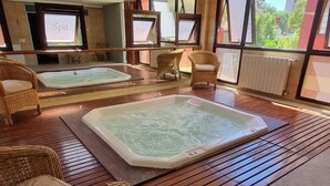 Área de spa com duas banheiras de hidromassagem, cadeiras de vime e uma grande janela com vista para prédios e árvores.
