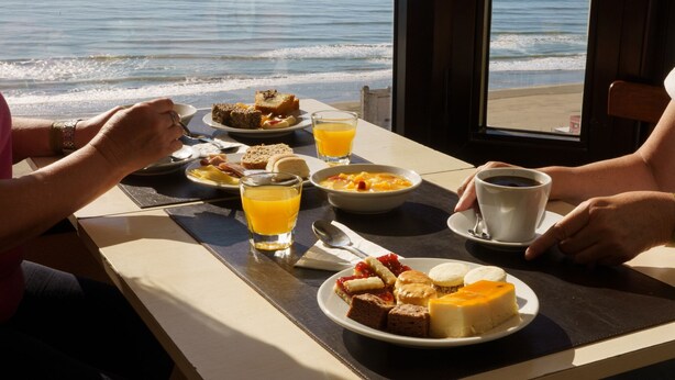 Mesa com itens de café da manhã, incluindo torradas, sopa e uma xícara de café, com vista para uma praia.