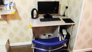 Quarto de canto com escrivaninha, computador, cadeira azul, chaleira e prateleira com objetos.