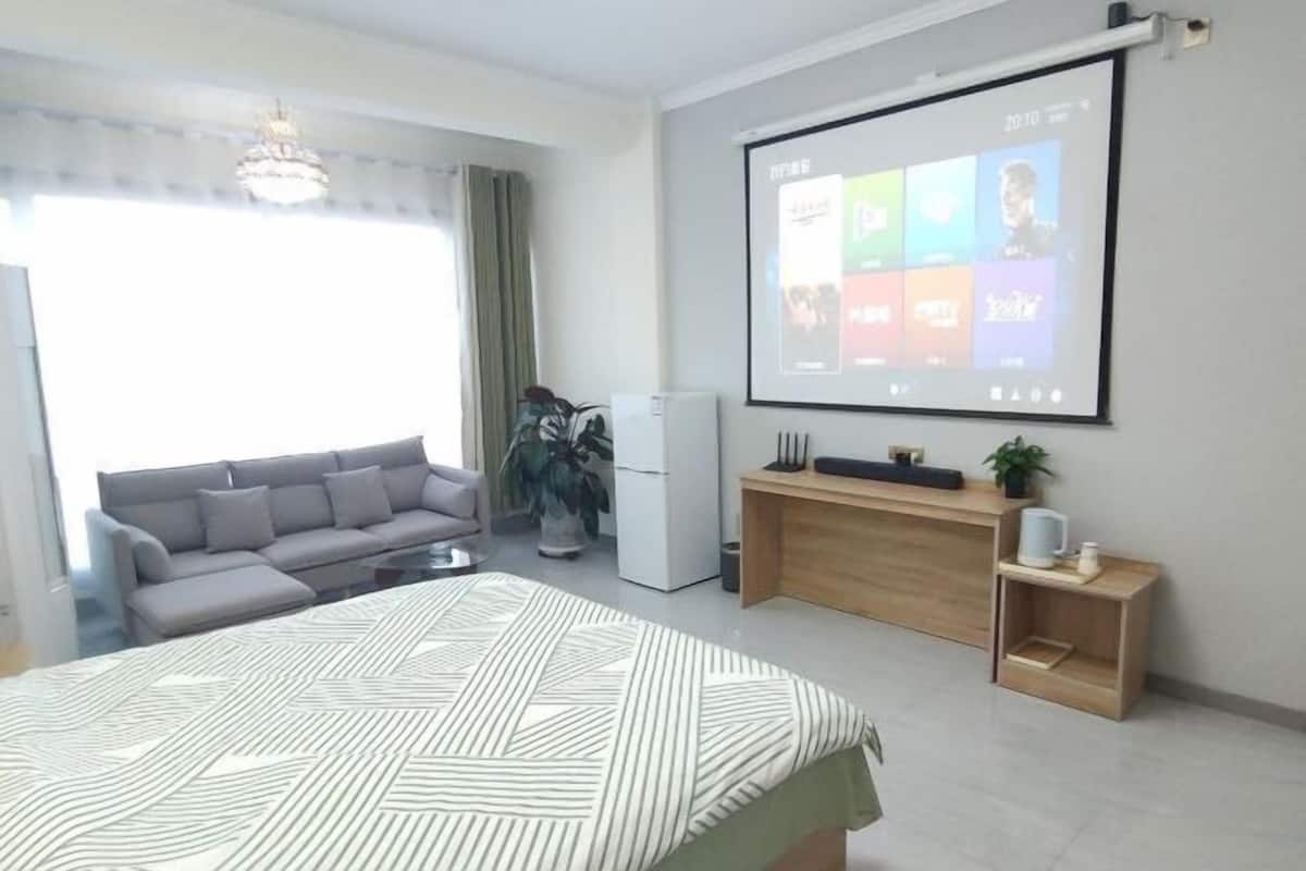 Quarto de hotel moderno com cama, sofá, escrivaninha, tela de projetor e geladeira.