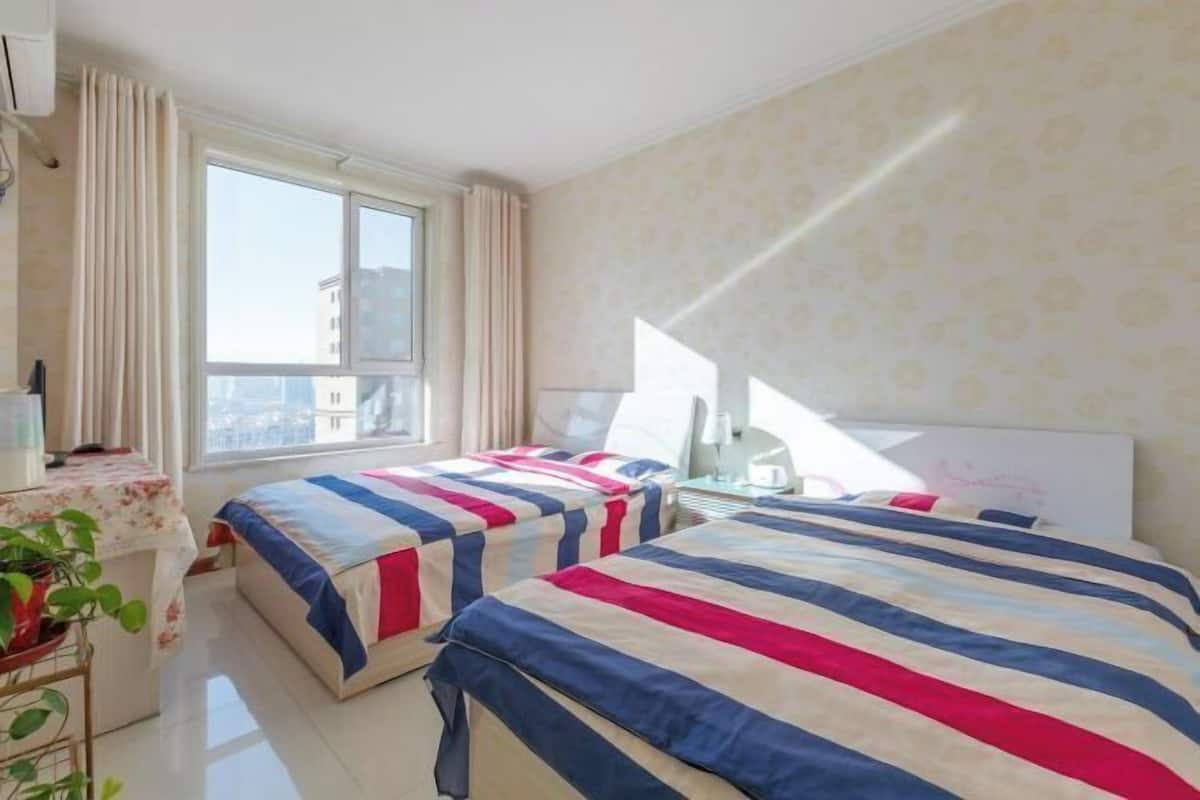 Quarto de hotel com duas camas, uma escrivaninha, uma cadeira, uma planta em vaso e uma janela grande.