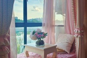 Quarto com uma grande janela, uma mesa de cabeceira com um vaso de flores, uma cama com manta rosa e vista para a cidade.