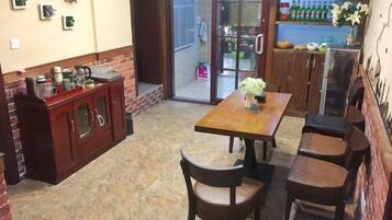 Uma pequena sala de jantar com mesa e cadeiras de madeira, um armário com utensílios de cozinha e vista para área externa através de uma porta de vidro.