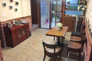 Uma pequena sala de jantar com mesa e cadeiras de madeira, um armário com utensílios de cozinha e vista para área externa através de uma porta de vidro.