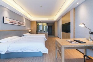 Quarto de hotel moderno com uma cama grande, uma escrivaninha, uma televisão e um banheiro visível por uma porta de vidro.