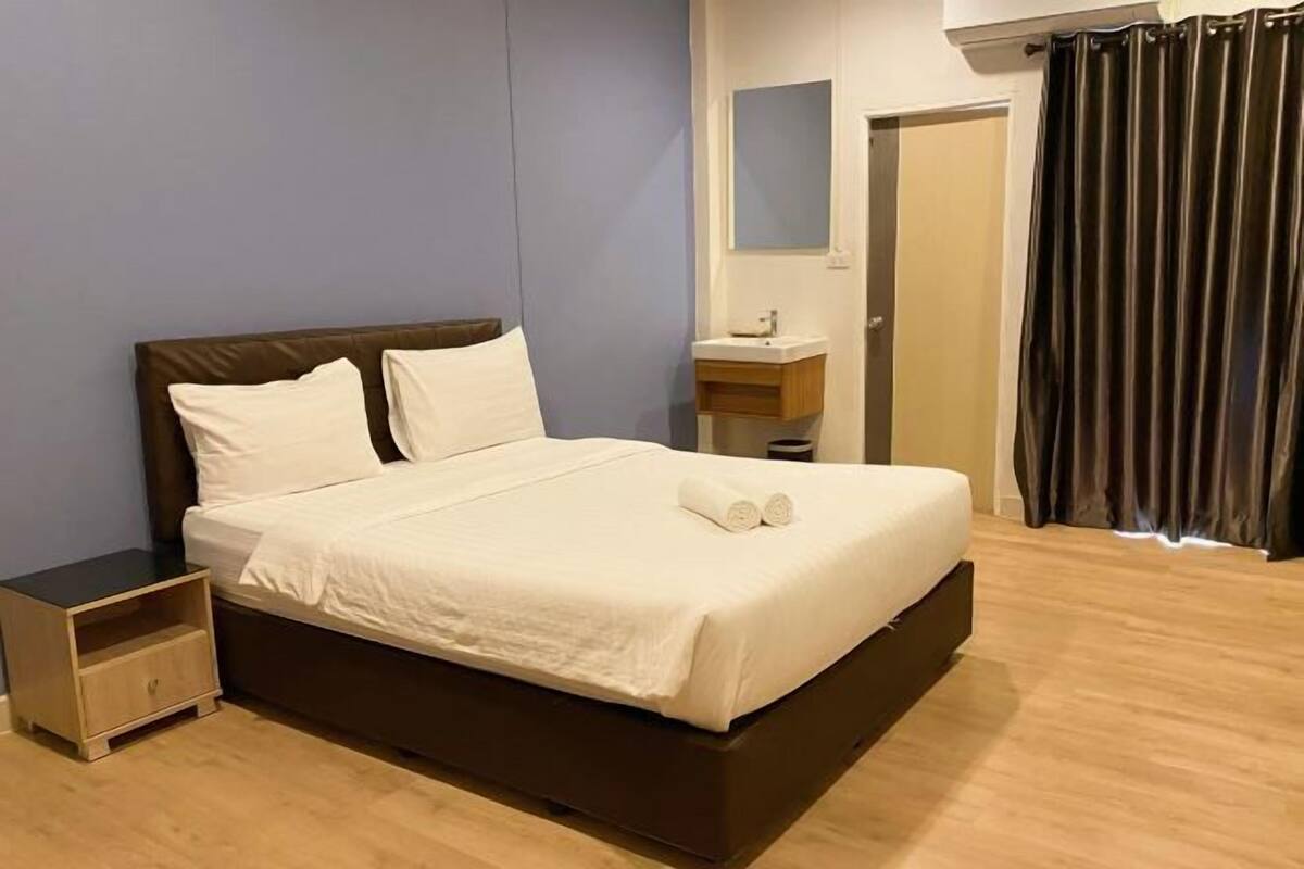 Cama bem arrumada com lençóis brancos e duas toalhas enroladas, uma mesa de cabeceira, um criado-mudo de madeira, um banheiro com pia e espelho, e uma cortina escura.