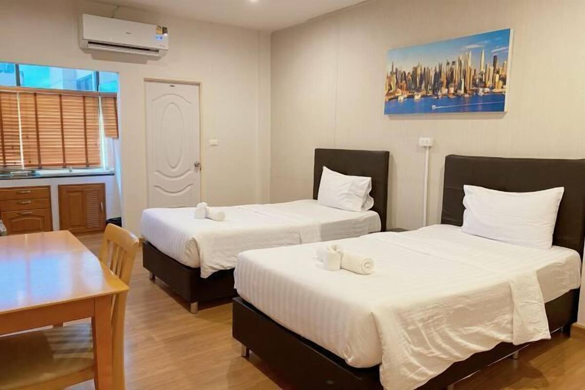 Quarto de hotel com duas camas, uma mesa de jantar, janela com persianas, ar condicionado e uma pintura de paisagem urbana na parede.