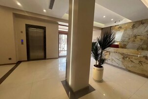Um hall de hotel com uma recepção de mármore, uma planta em vaso e um elevador.