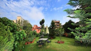 Um jardim bem cuidado com área de refeições, rodeado por vegetação exuberante e edifícios ao fundo.