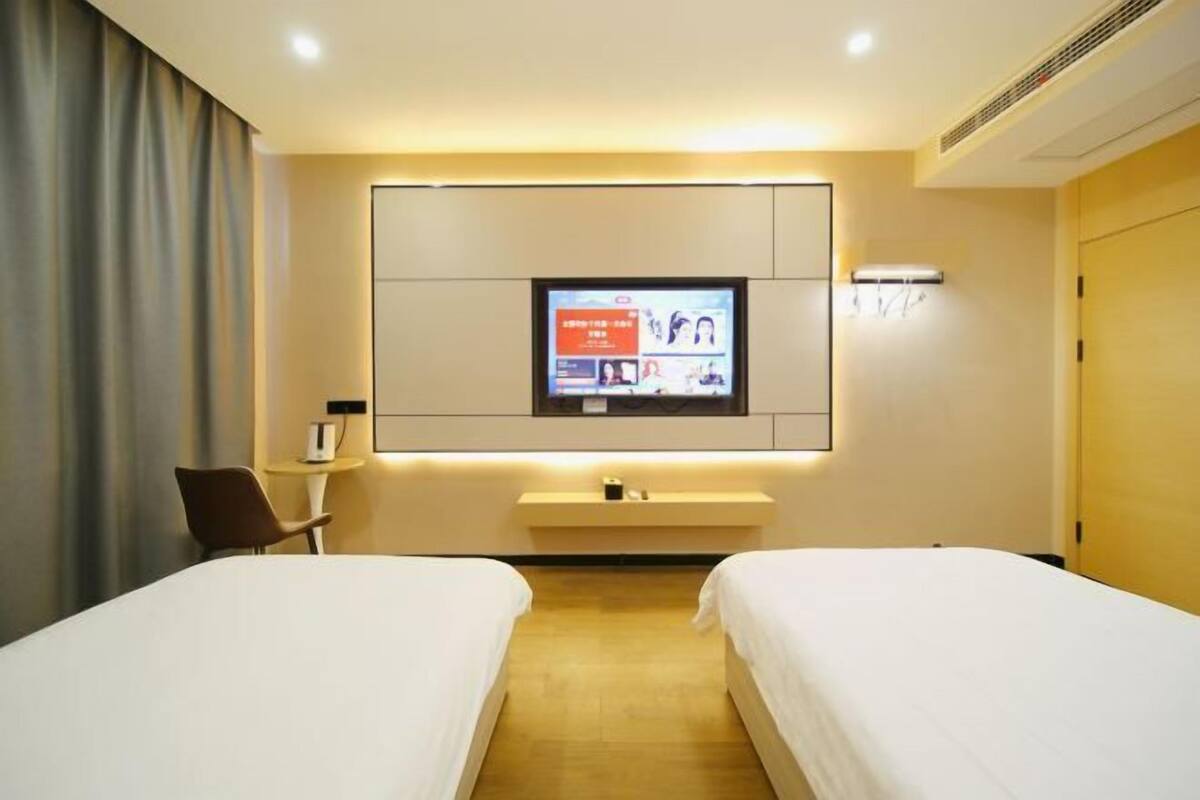 Quarto de hotel com duas camas, uma televisão na parede, uma pequena mesa com uma cadeira e uma mesa de cabeceira com um abajur.