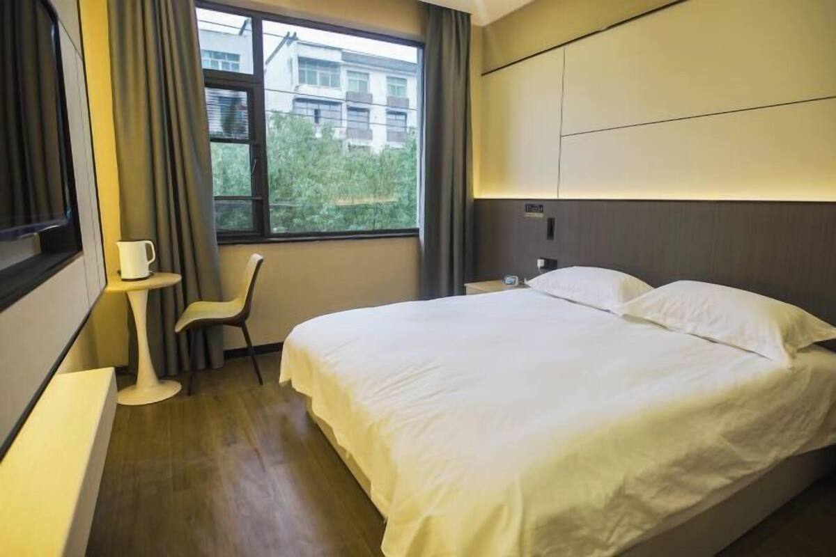 Quarto de hotel com uma cama grande, uma mesa de cabeceira, uma cadeira, uma janela com cortinas e vista para edifícios.