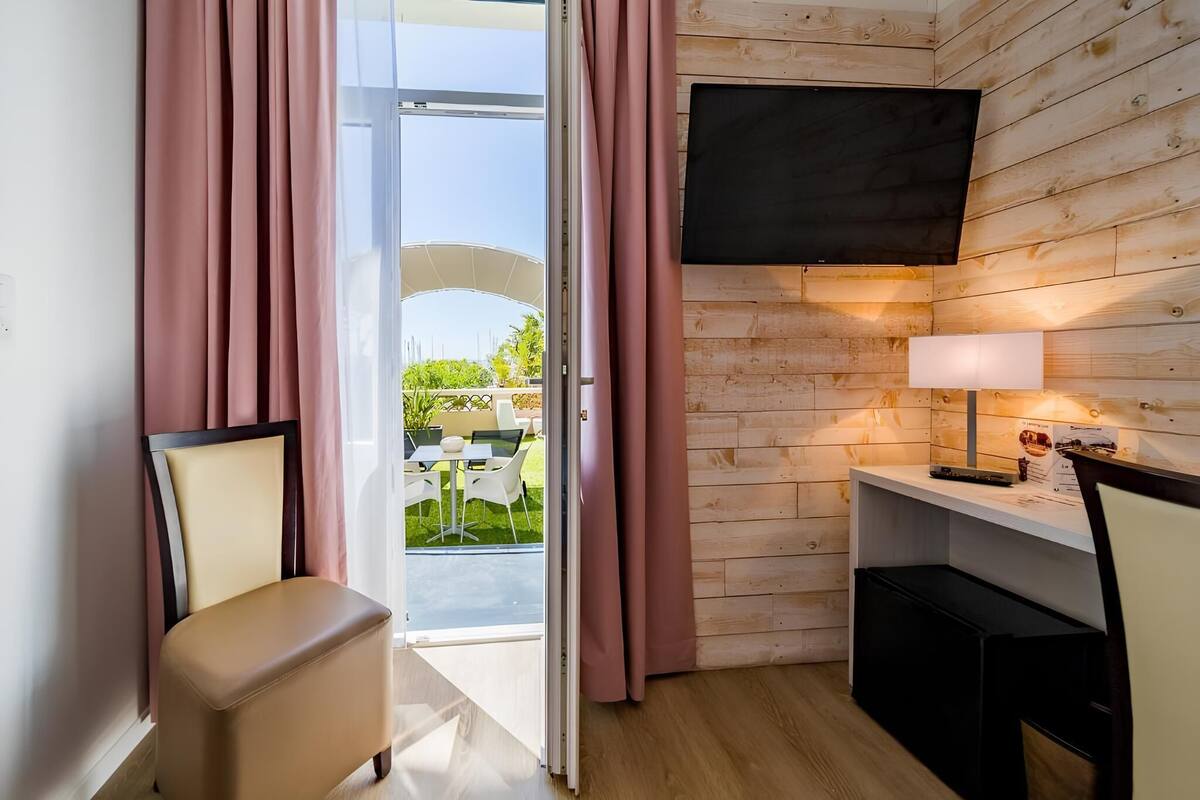 Quarto com porta de vidro deslizante, televisão de tela plana na parede, uma escrivaninha pequena com abajur e uma cadeira.