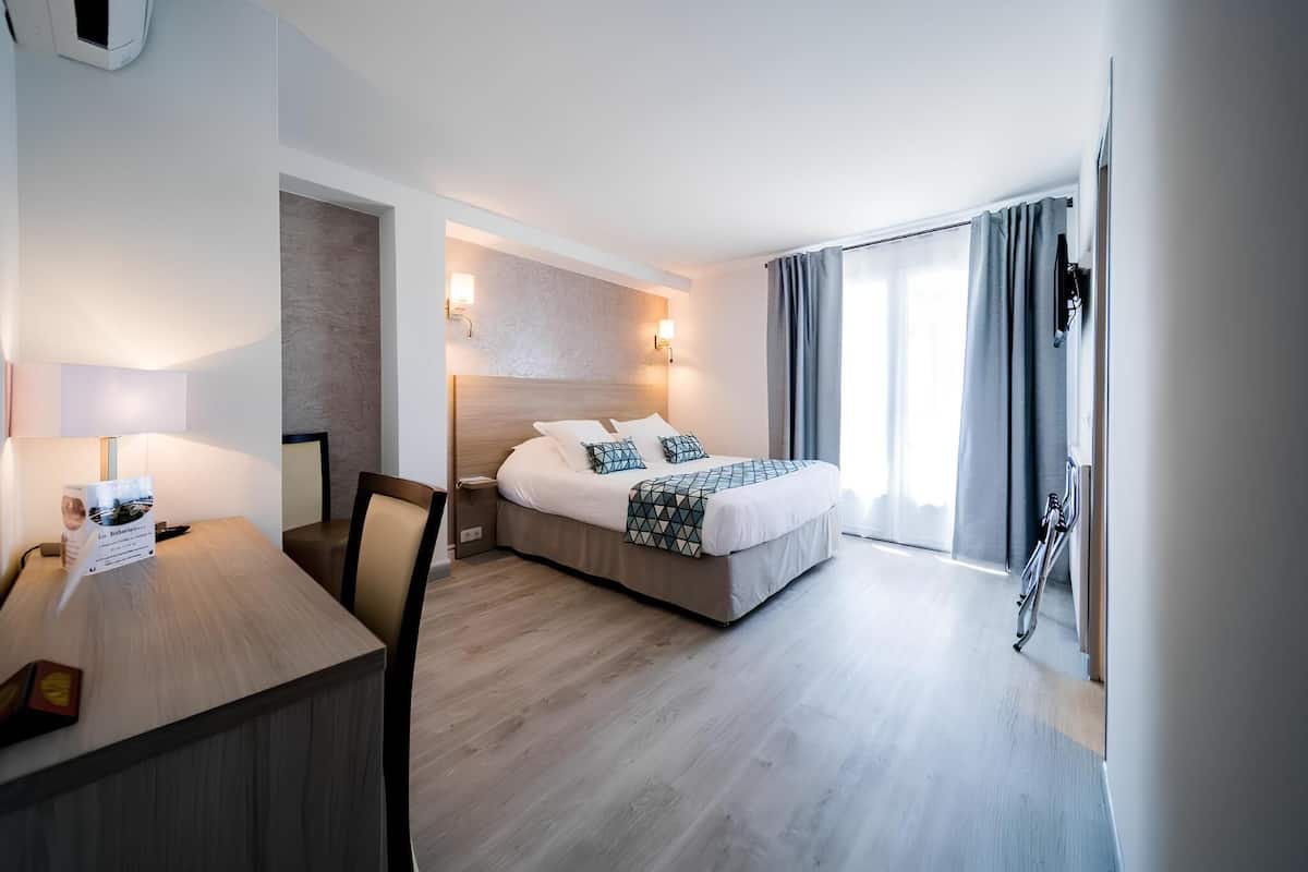 Quarto de hotel moderno com uma cama, uma escrivaninha e uma cadeira. Há uma janela com cortinas e vista para o exterior.