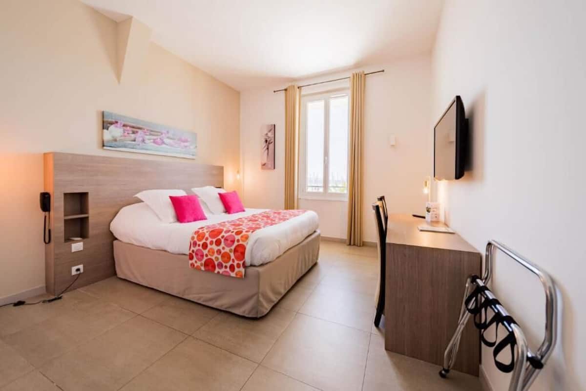 Quarto de hotel com cama, mesa de cabeceira, escrivaninha, cadeira e janela com cortinas.