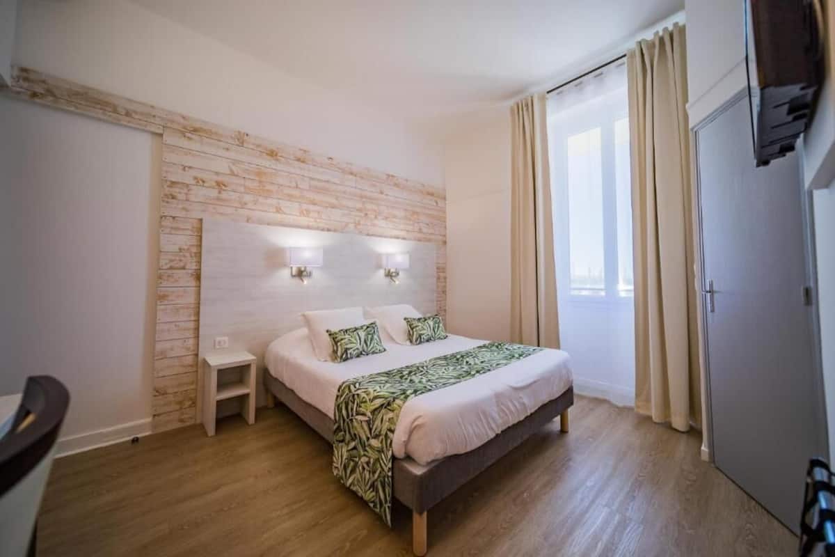 Quarto com cabeceira de madeira, cama com manta de padrão de folhas verdes, mesa de cabeceira e janela com cortinas.