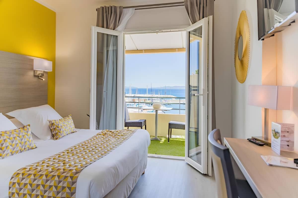 Quarto de hotel com cama, escrivaninha, cadeira, varanda com vista para o mar e televisão na parede.