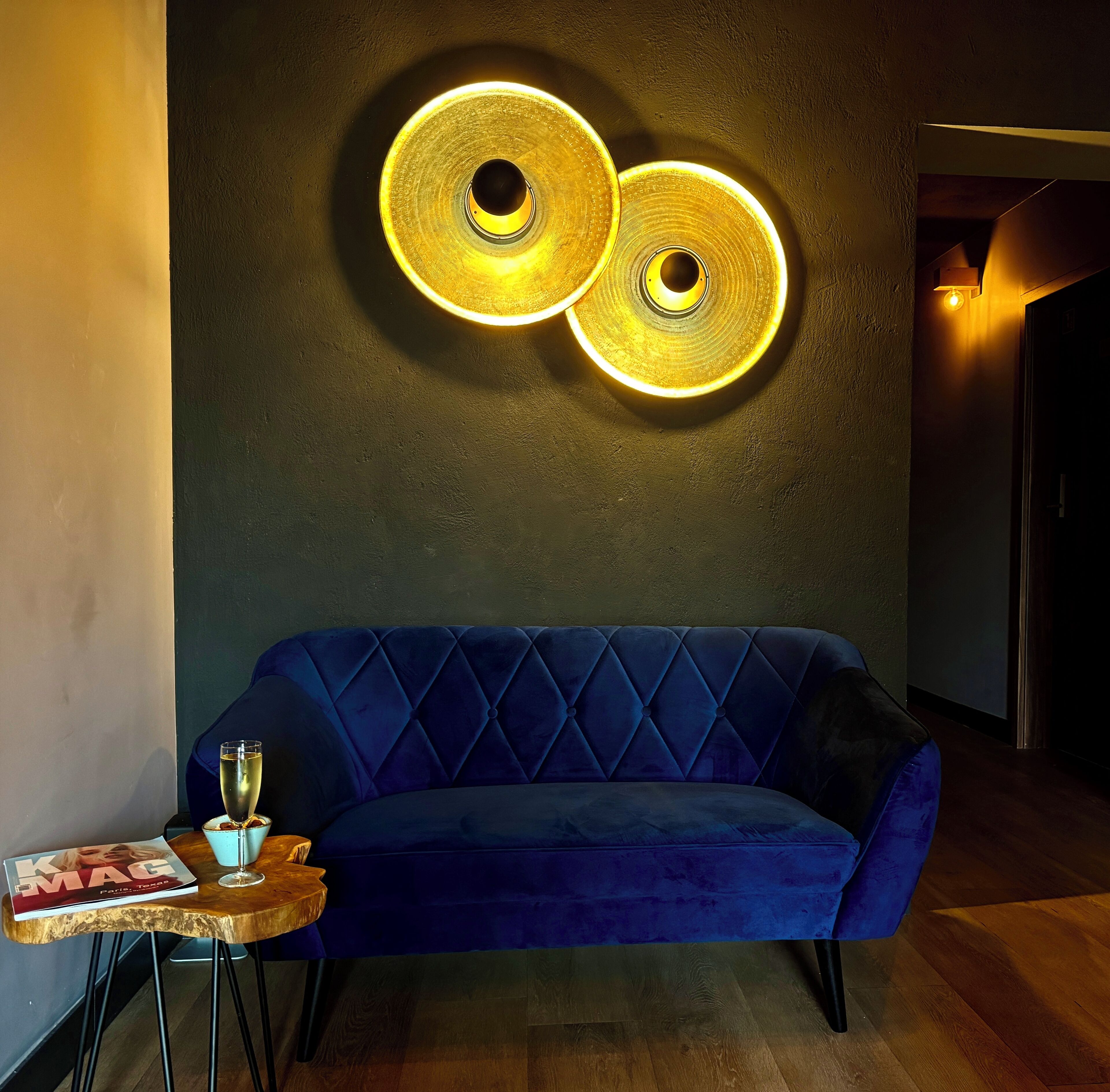 Um quarto com um sofá de veludo azul, uma pequena mesa redonda com uma taça de champanhe e duas luminárias circulares fixadas na parede.