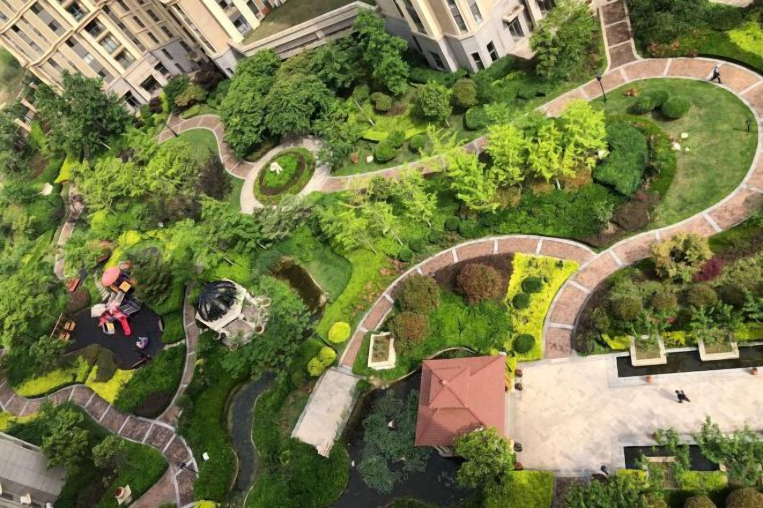 Vista aérea de um pátio ajardinado com caminhos, uma fonte central e vegetação ao redor.