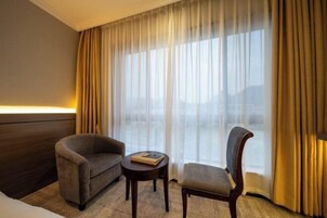 Quarto de hotel com cama, sofá, poltrona, mesa pequena e uma janela grande com cortinas.
