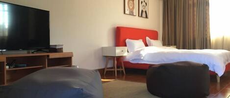 Quarto de hotel com televisão de tela plana, cama com cabeceira vermelha, duas fotos emolduradas na parede, um pufe grande e uma escrivaninha de madeira.