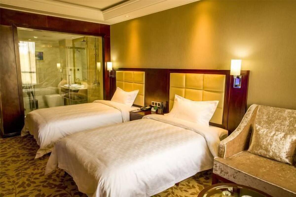 Quarto de hotel com duas camas, um sofá, cabine de hidromassagem em vidro e cabeceira de madeira.