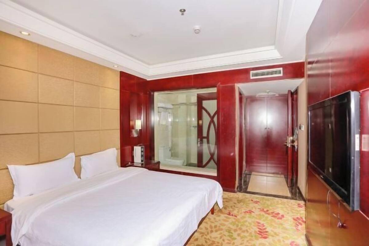 Quarto de hotel com uma cama grande, televisão, banheiro com porta de vidro e uma parede vermelha decorativa.