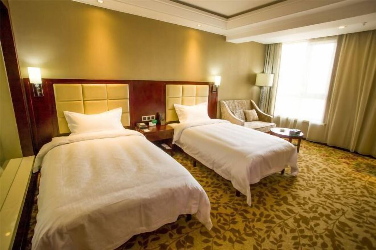 Quarto de hotel com duas camas, uma cadeira, uma mesinha e uma janela com cortinas.