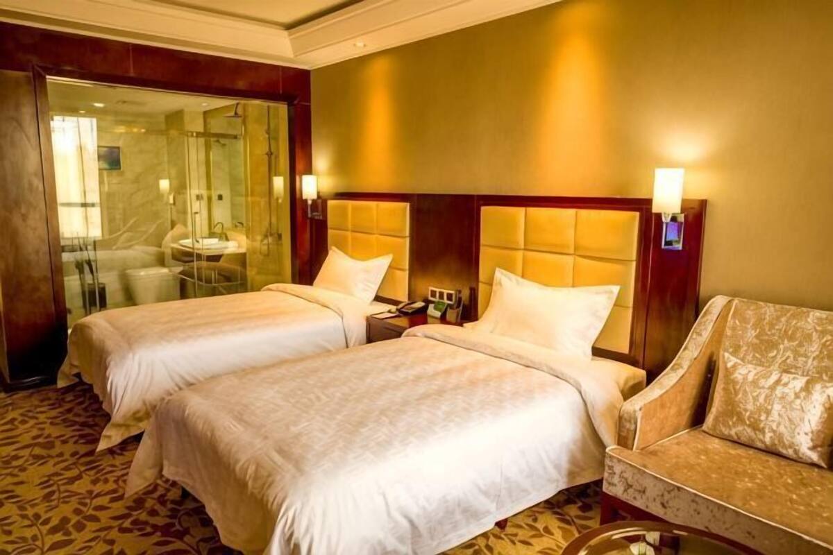 Quarto de hotel com duas camas, um sofá, área de chuveiro com divisória de vidro e uma pequena mesa com luminária.